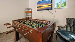 Um quarto com mesa de pebolim, televisão na parede e janela com vista para árvores.