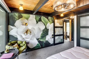 Um interior moderno com um grande mural floral, uma poltrona verde, uma mesa lateral de vime com um livro e uma cama com cabeceira branca.