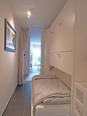 Corredor de hotel estreito, com uma cama à direita, um quadro emoldurado à esquerda e uma porta que dá para um quarto iluminado.