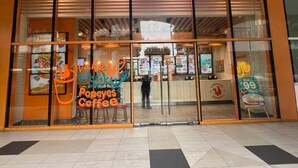 Um restaurante Popeyes com uma grande janela de vidro, um letreiro anunciando 'Popeyes Coffee' e um quadro de menu visível através da janela.