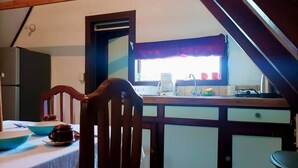Uma área de refeições com uma mesa posta para dois, uma cadeira e vista para uma cozinha com janela.