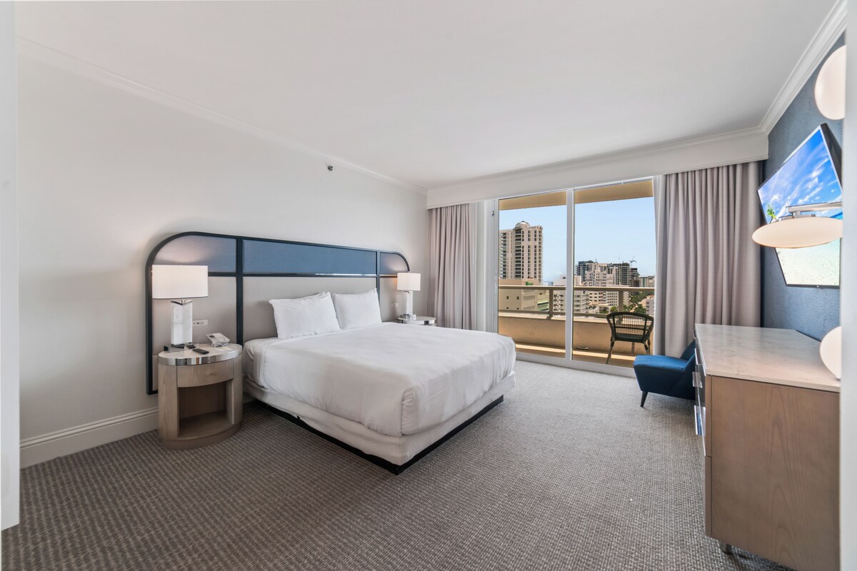Quarto de hotel com cama grande, escrivaninha, cadeira e vista da cidade pela janela.