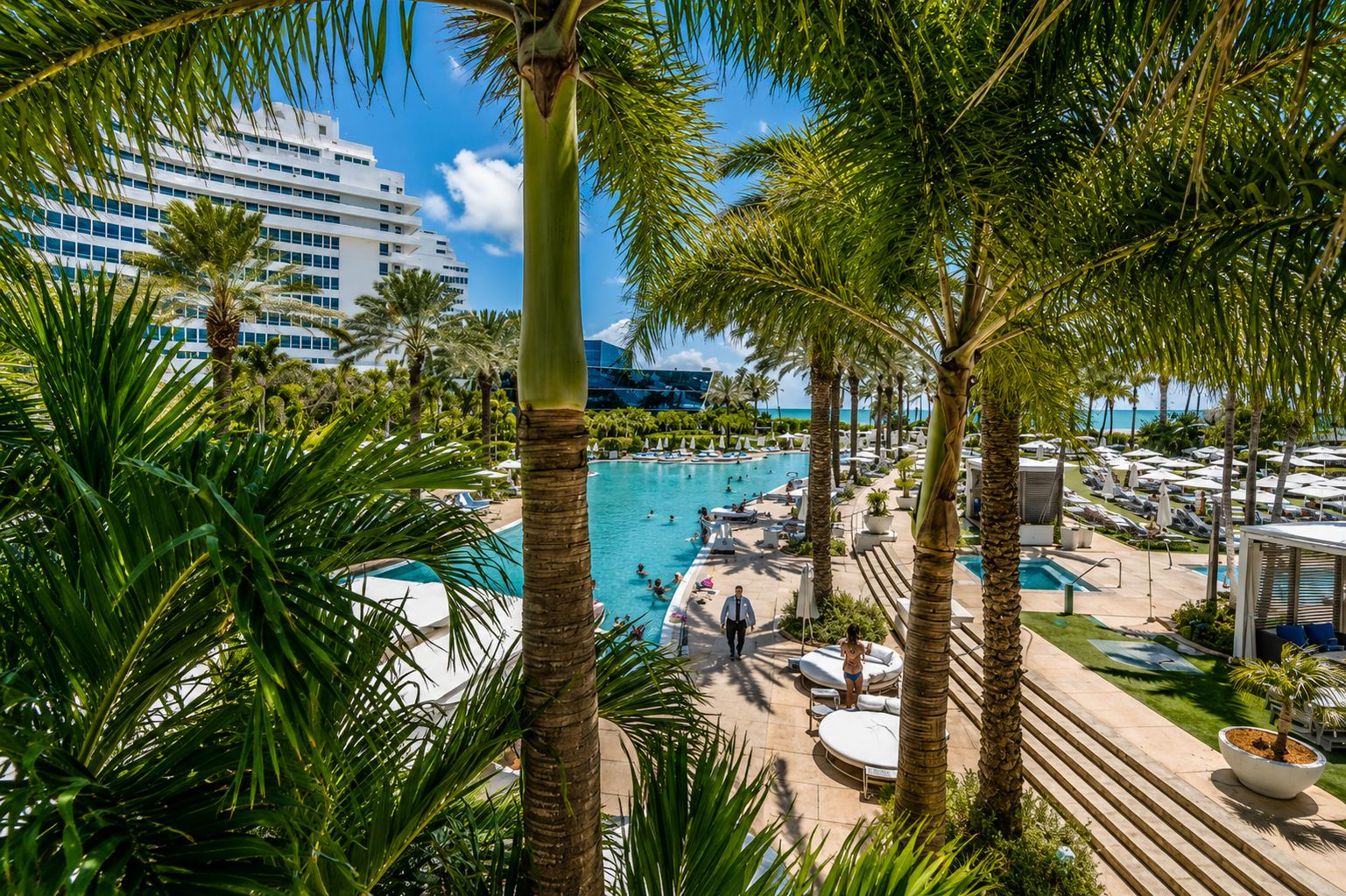 Área de piscina em resort, com palmeiras, espreguiçadeiras e um edifício grande ao fundo.