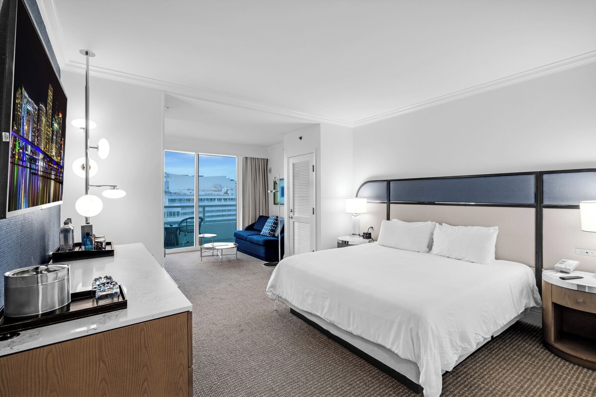 Quarto de hotel com uma cama grande, uma mesa com luminária, uma cadeira e vista para a cidade pela janela.