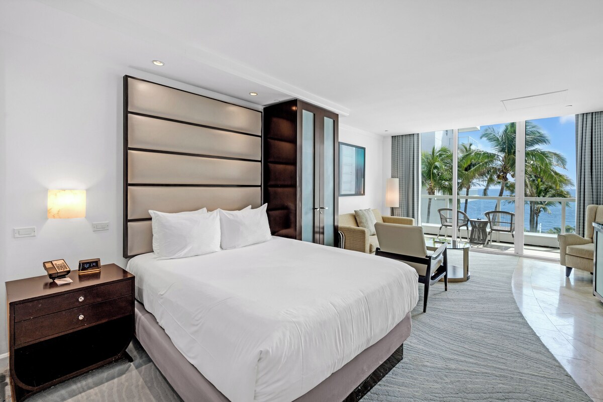 Quarto de hotel moderno com uma cama grande, uma mesa de cabeceira, uma escrivaninha e vista para o oceano.