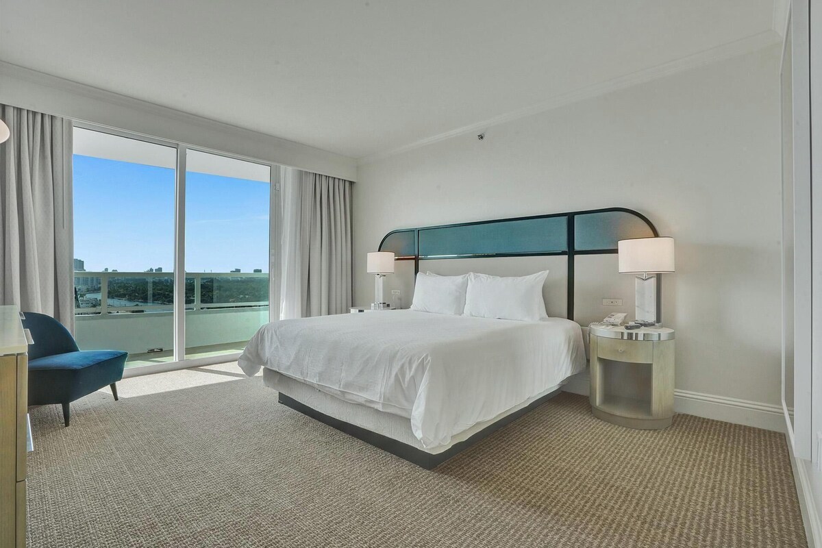 Quarto de hotel com uma cama grande, duas mesas de cabeceira com abajures, uma cadeira e vista para a paisagem urbana através de grandes janelas.