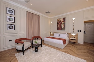 Quarto de hotel com cama, duas cadeiras, uma mesinha, um quadro e uma porta.