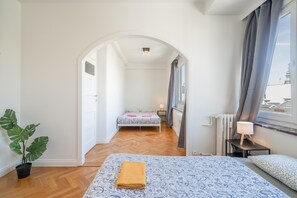 Quarto de hotel com cama, mesa de cabeceira, abajur, planta em vaso e vista da cidade pela janela.