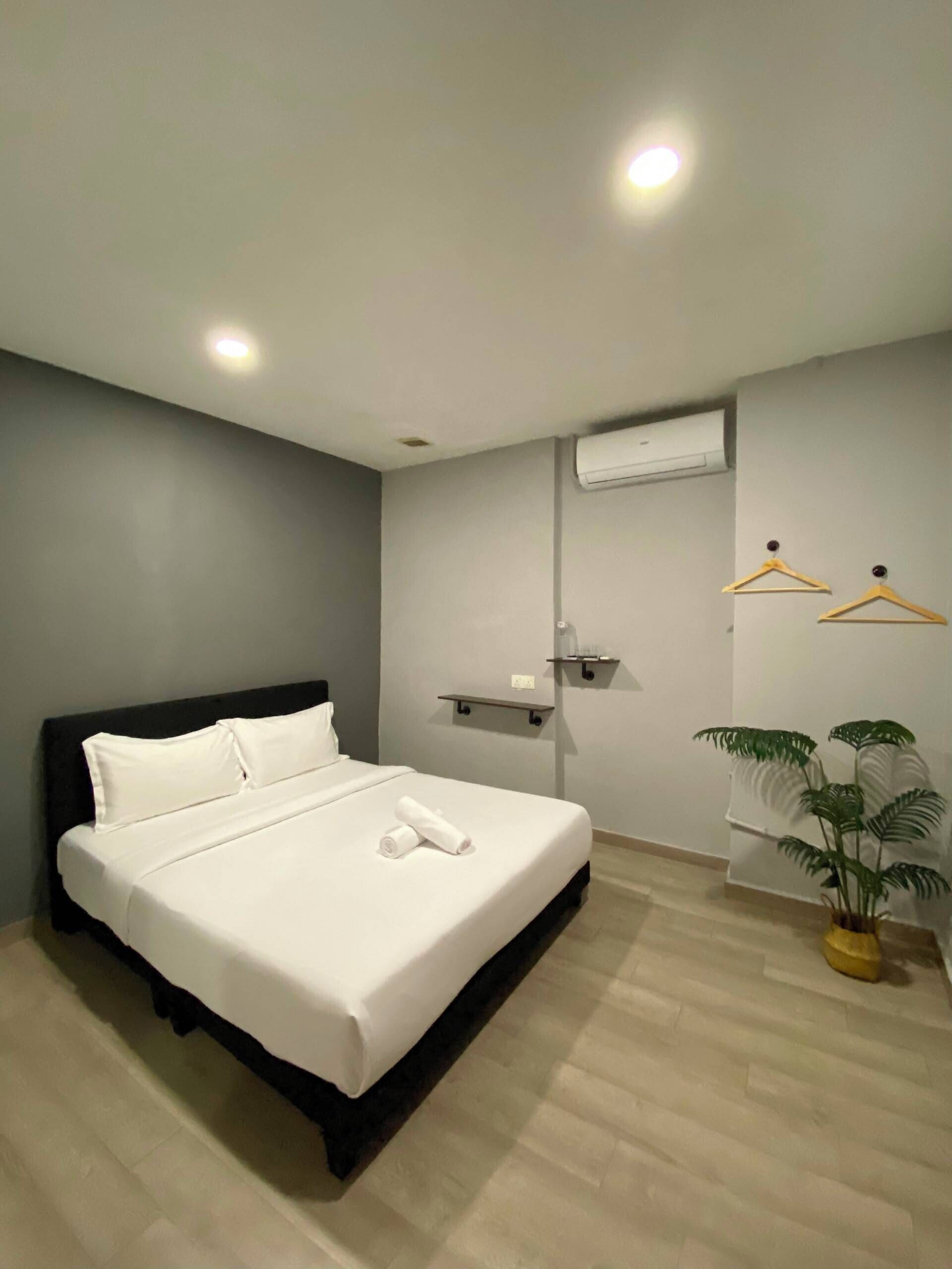 Quarto de hotel moderno com uma cama grande, uma mesa de cabeceira, um aparelho de ar condicionado, uma pequena prateleira e um vaso de plantas.