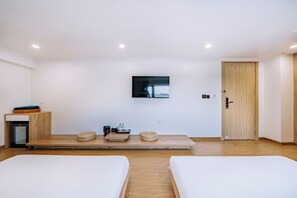 Quarto de hotel com duas camas, uma televisão, uma mesinha com uma cesta, um cesto de lixo e uma porta de madeira.