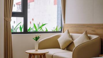 Quarto com um sofá bege, uma pequena mesa de madeira e janela com vista para vegetação e edifícios.