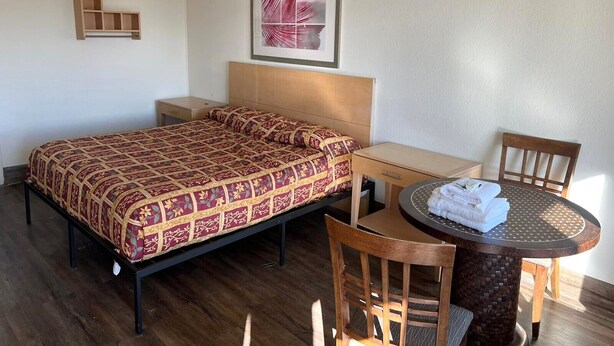 Quarto de hotel com uma cama, uma mesinha com cadeiras, criado-mudo e prateleira na parede.