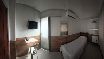 Quarto compacto com cama, uma pequena mesa, uma cadeira, TV fixada na parede e janela com cortinas.