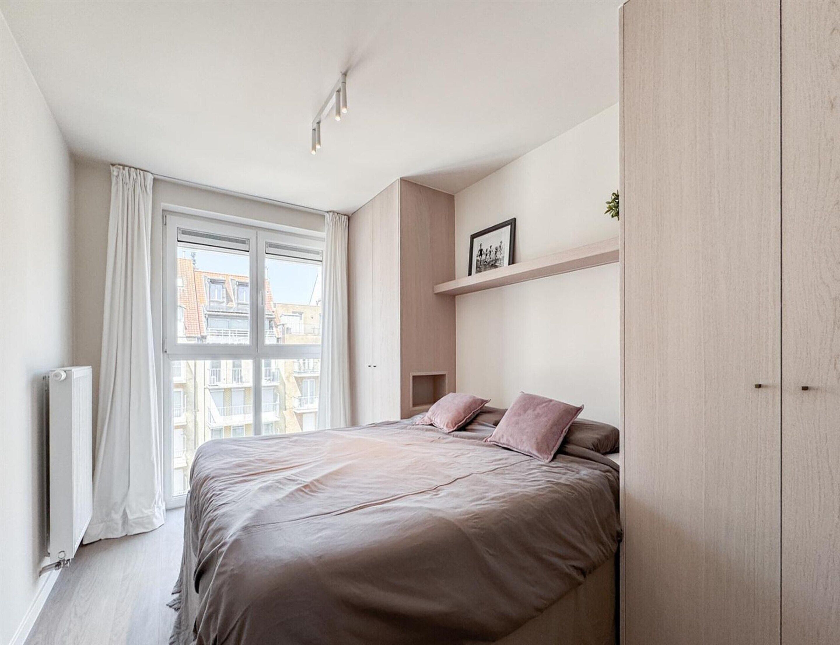 Quarto com cama, radiador, janela com cortinas, prateleira com um quadro e uma planta.