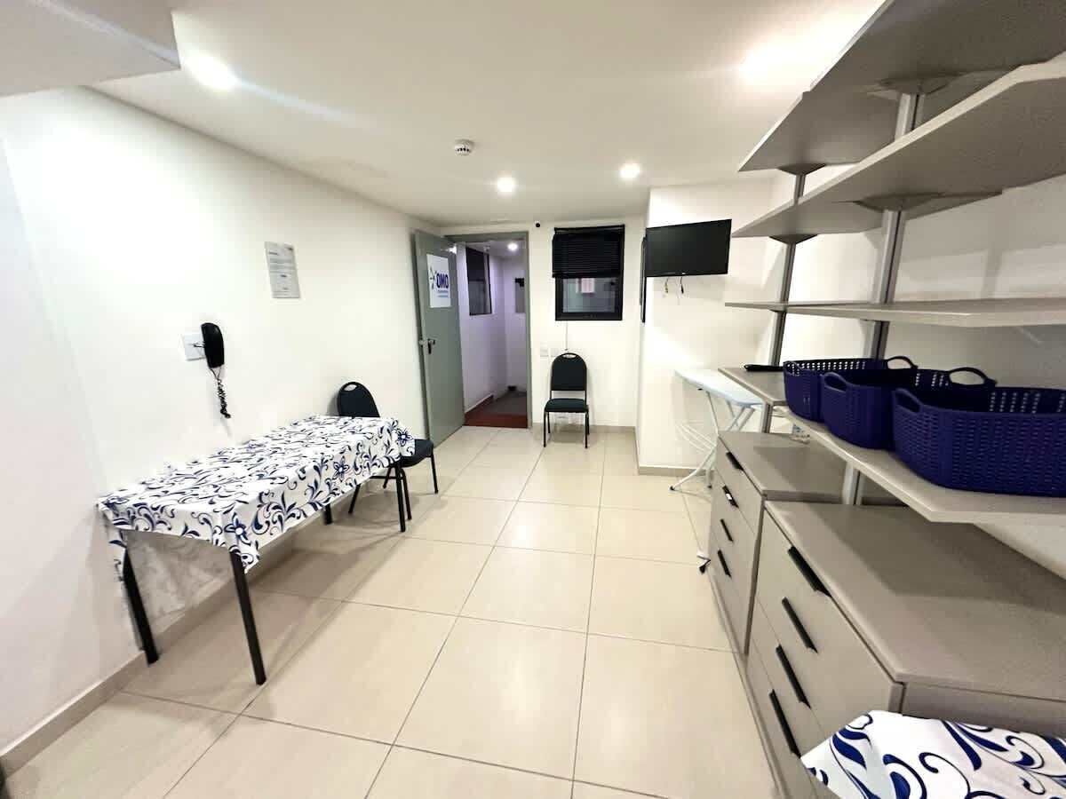 Um quarto com uma mesa comprida, cadeiras, uma escrivaninha com gavetas e estantes com cestos.