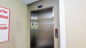 Um elevador com visor digital, botão de alarme de incêndio e sinalização indicando a localização dos corredores do primeiro andar e do jardim.