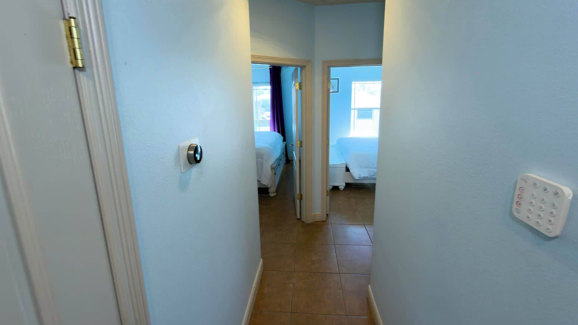 Corredor estreito com uma porta à esquerda, um interruptor de luz à direita e uma cama visível ao fundo.