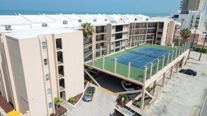 Um edifício de vários andares com quadra de tênis no térreo, cercado por estacionamento e vista para o mar.
