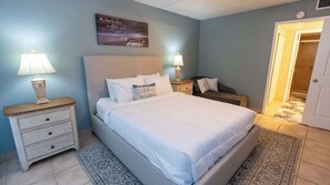 Quarto com uma cama, duas mesinhas de cabeceira, um criado-mudo, uma luminária na parede, uma porta e um tapete estampado.
