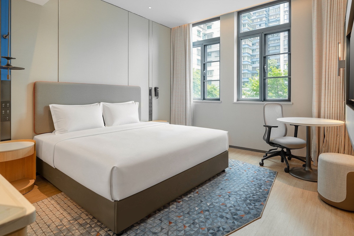 Quarto de hotel com cama, escrivaninha, cadeira, mesa pequena e uma janela grande com vista para prédios.