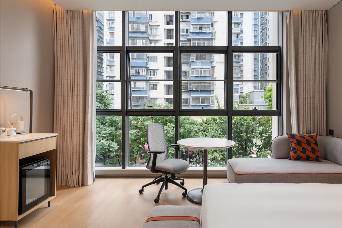 Quarto de hotel com cama, escrivaninha com luminária, cadeira, mesa pequena e vista para edifícios altos através de janelas amplas.