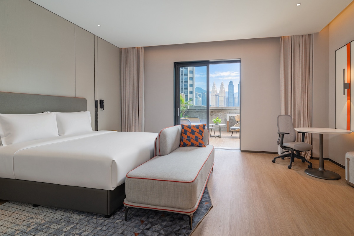 Quarto de hotel com uma cama grande, uma escrivaninha com cadeira, uma mesinha e uma televisão.