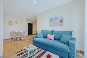 Uma sala de estar com um sofá azul, uma mesa de jantar com cadeiras, um tapete colorido e uma pintura com tema de praia na parede.