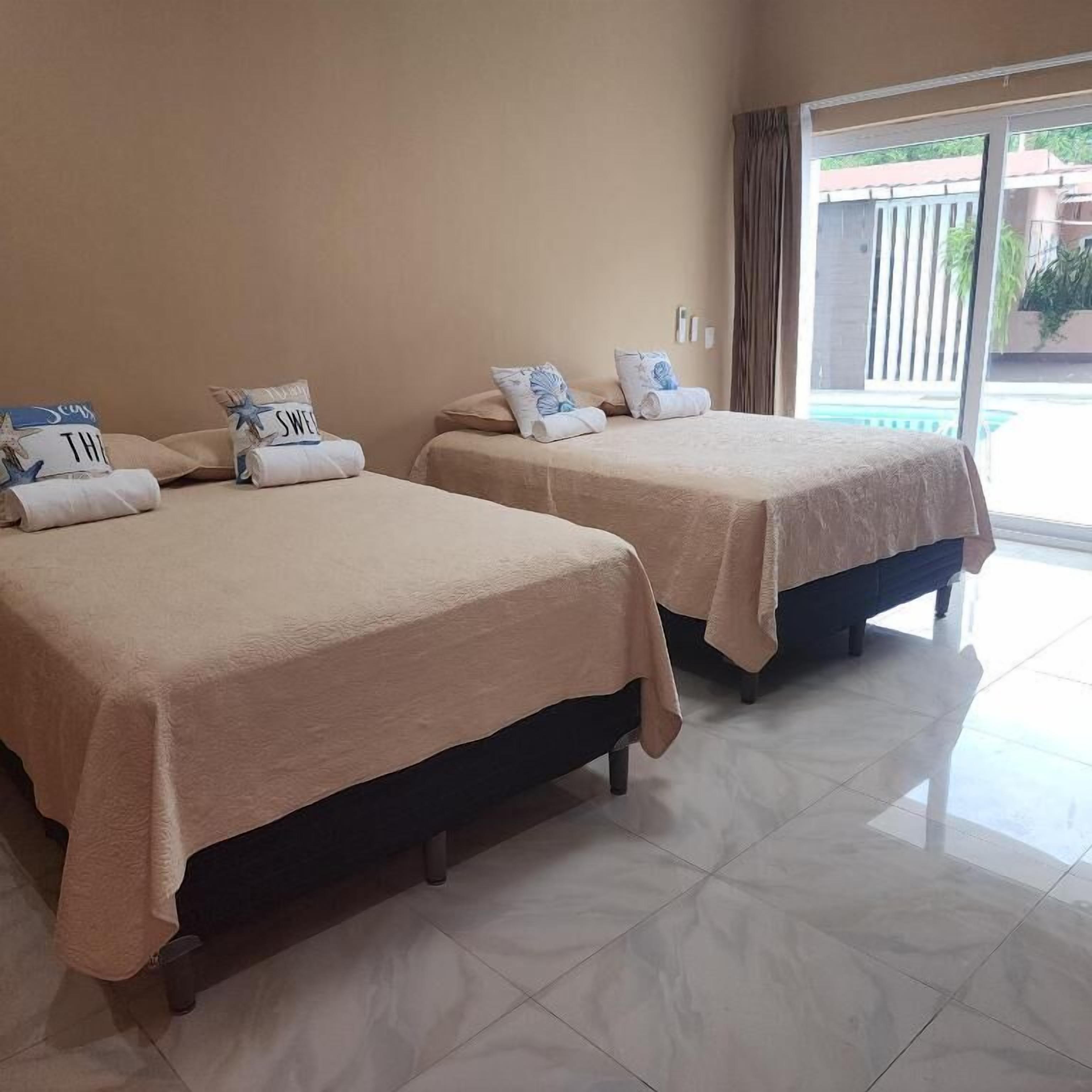 Quarto de hotel com duas camas, cada uma com colcha bege e almofadas com texto.
