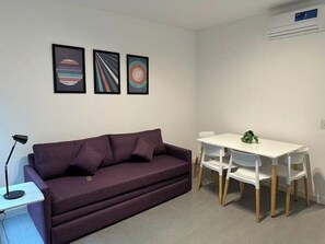 Uma sala moderna com um sofá roxo, uma mesa de jantar branca com cadeiras e três obras de arte abstratas nas paredes.