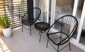 Varanda com duas cadeiras de vime preto e uma mesa redonda pequena, um vaso de planta e uma cama com roupa de cama e almofadas brancas.