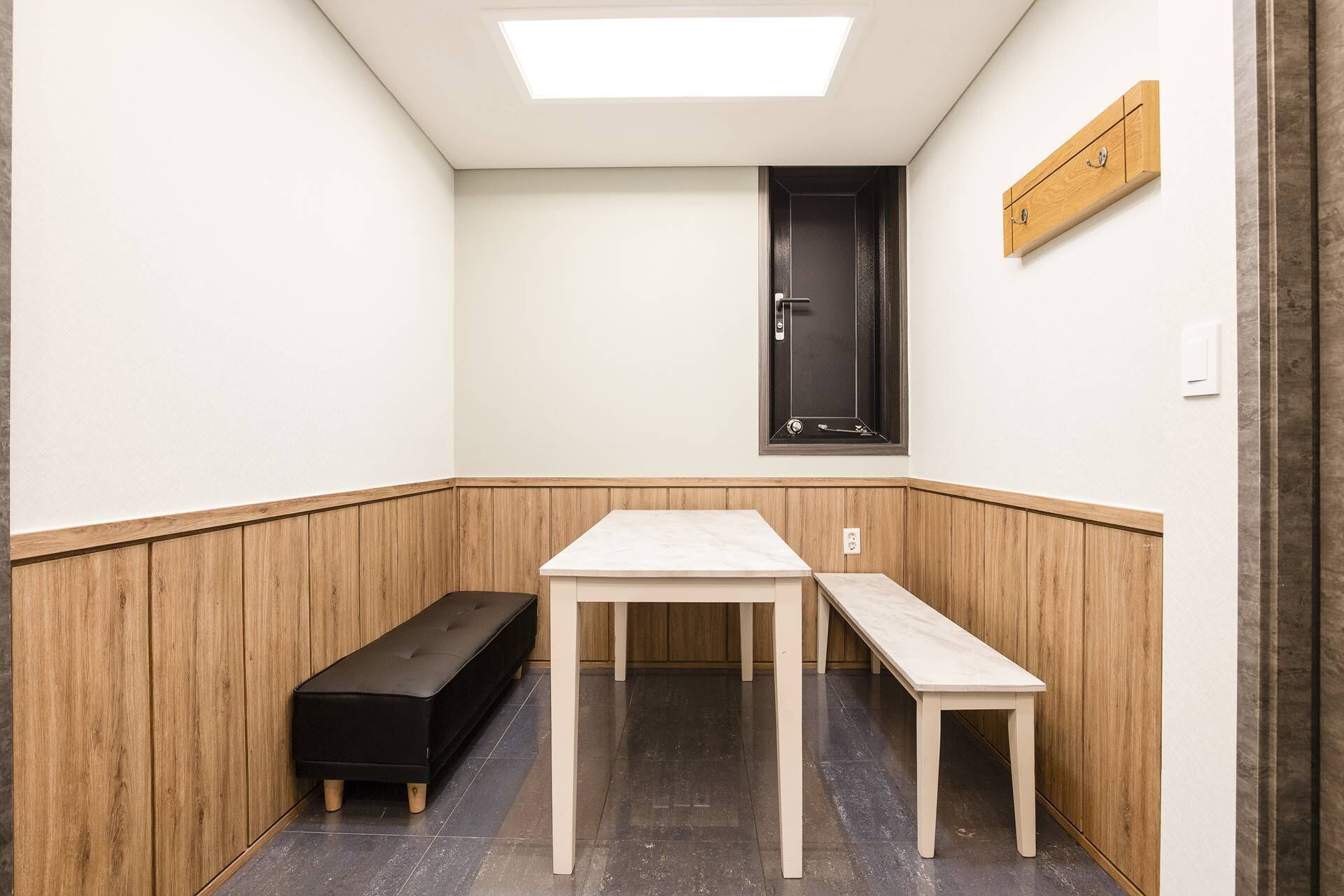 Um quarto pequeno e estreito, com um banco de madeira, uma mesa e uma porta.
