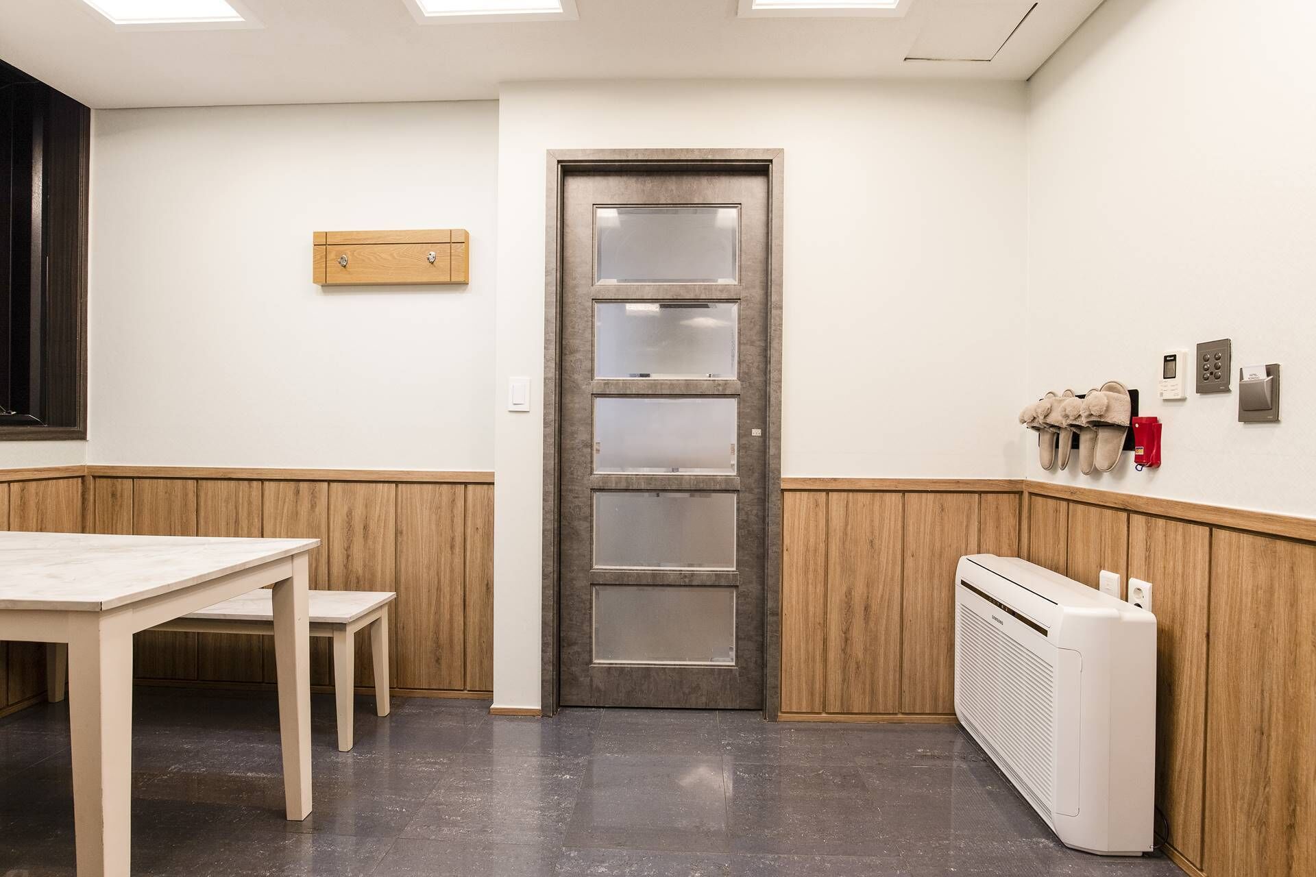 Um quarto com uma mesa de madeira, um banco, uma porta com painéis de vidro fosco, um aquecedor fixado na parede e um termostato na parede.