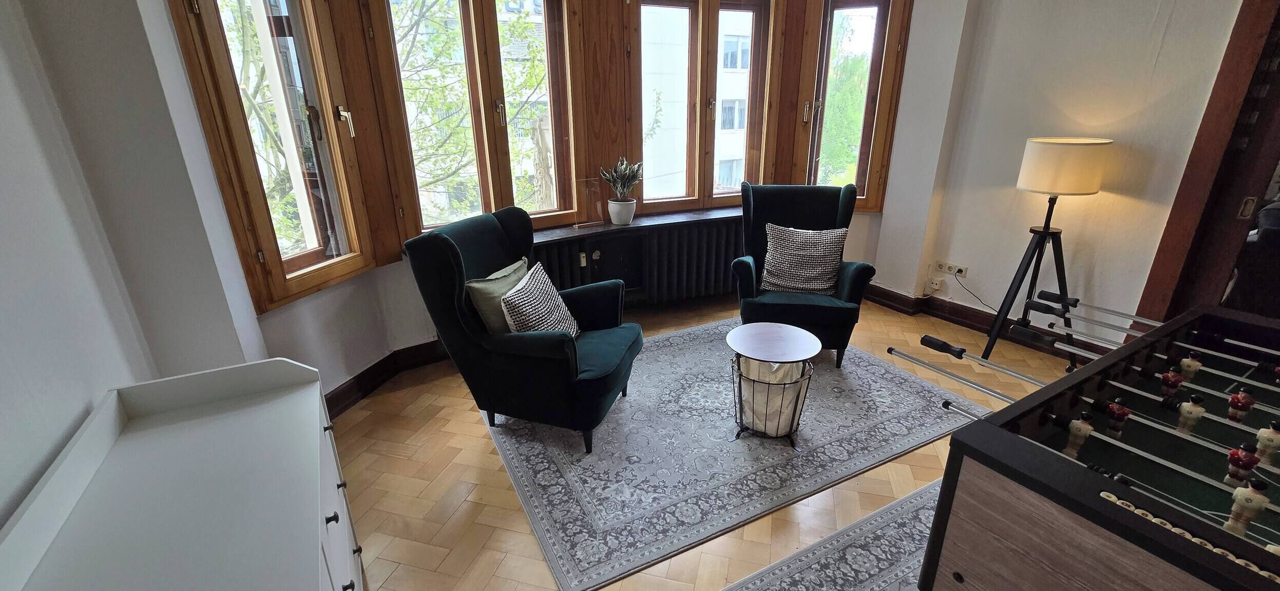 Um quarto com duas poltronas, uma mesinha redonda, um abajur e uma mesa de pebolim.