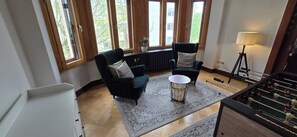 Um quarto com duas poltronas, uma mesinha redonda, um abajur e uma mesa de pebolim.