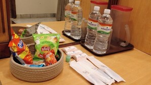 Uma área de recepção de hotel com um balcão de madeira, uma cesta de palha com lanches, água engarrafada e uma caixa de lenços.