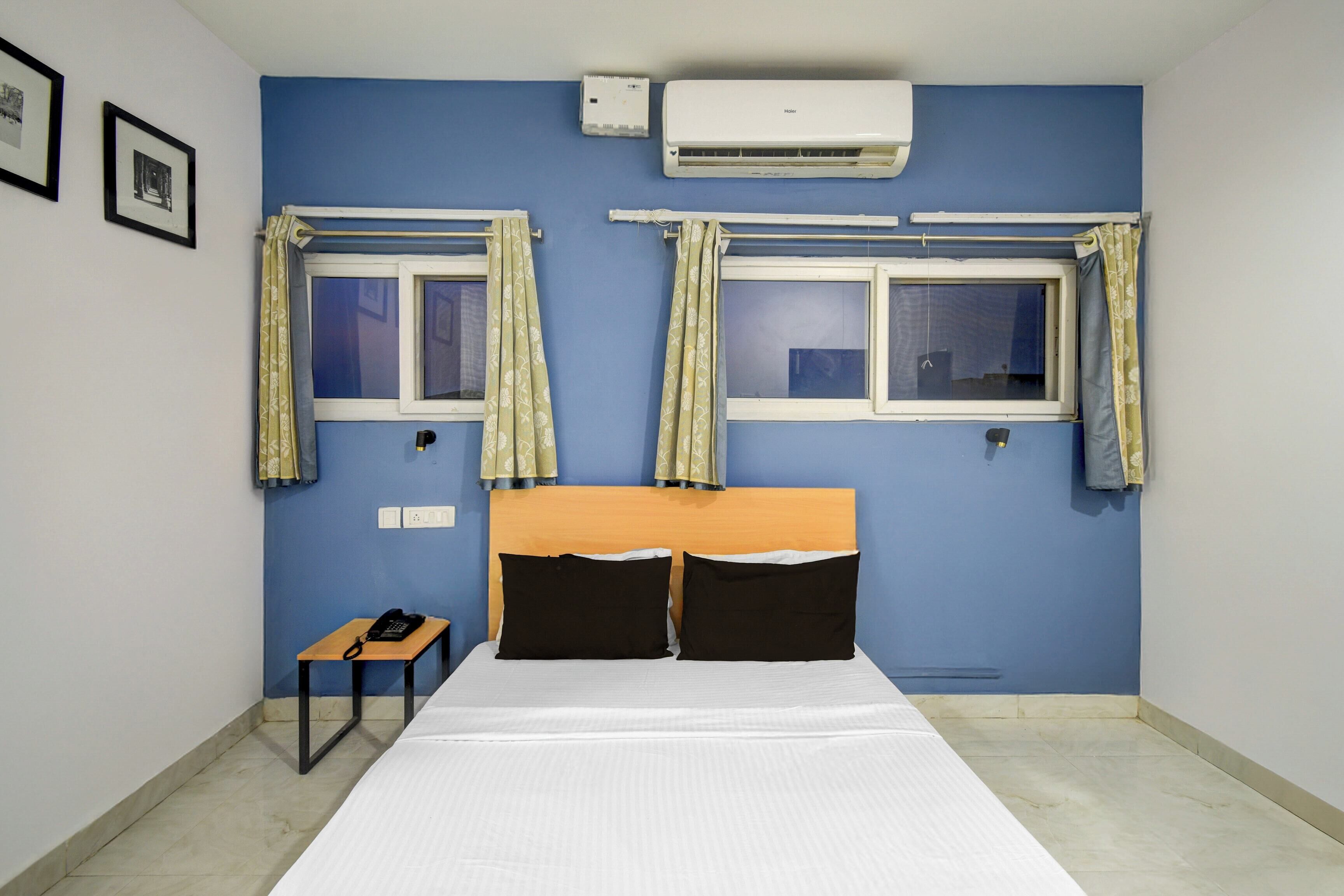 Quarto com duas janelas, uma cama, mesa de cabeceira com telefone e ar condicionado na parede.