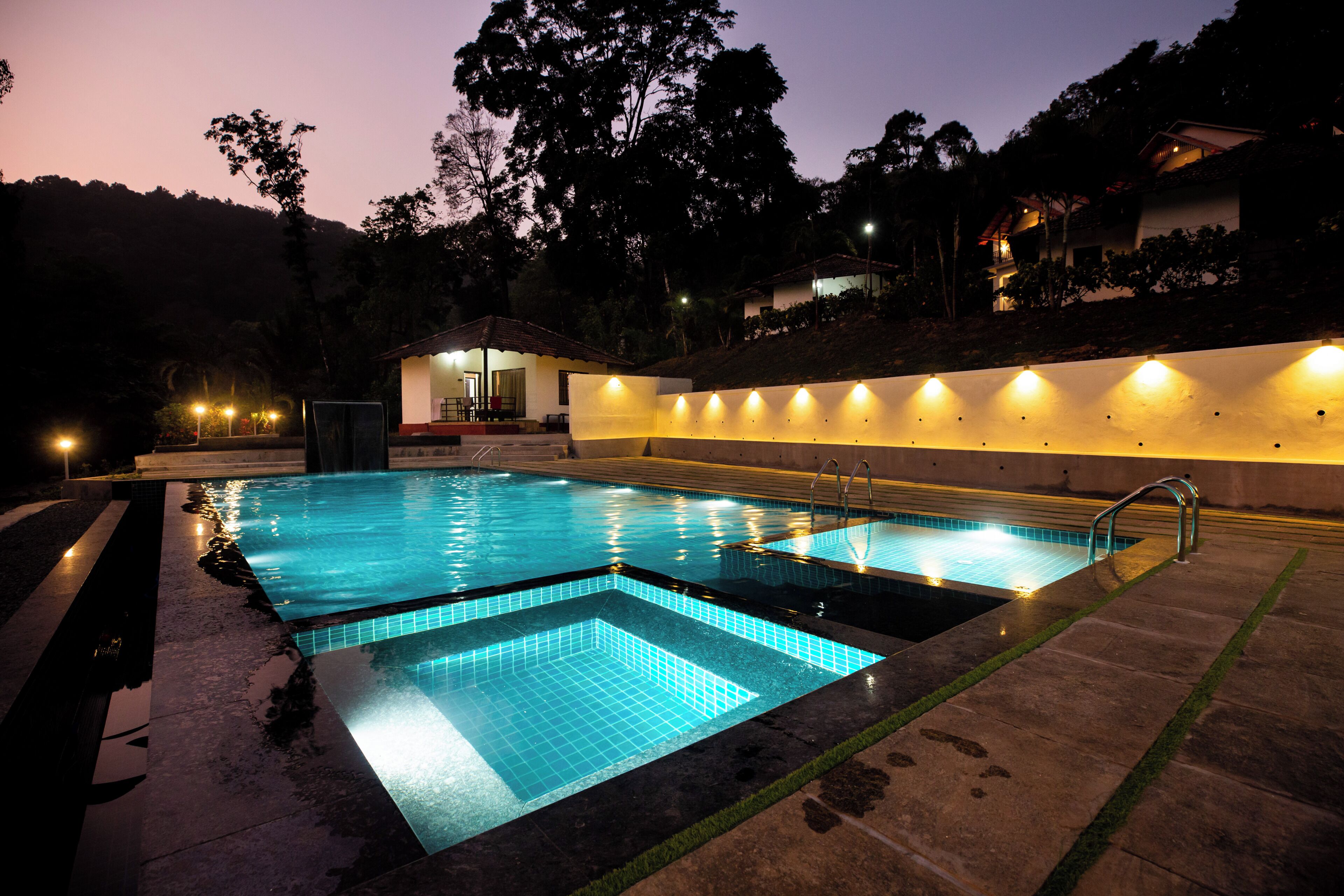 Uma piscina com borda azulejada, uma pequena estrutura adjacente e um muro com iluminação.