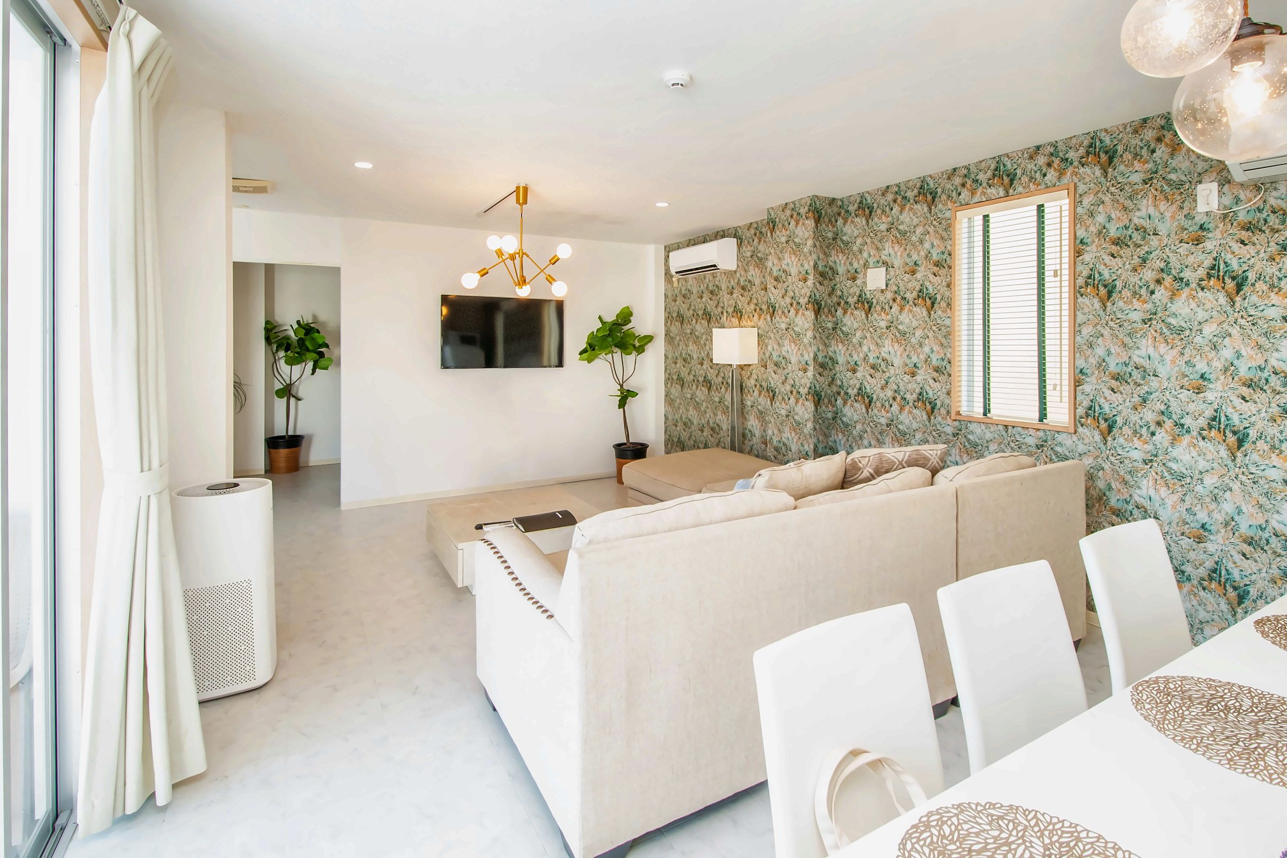 Uma sala de estar moderna com sofá, mesa de jantar e cadeiras. Há uma TV fixada na parede, um lustre e um ar condicionado.