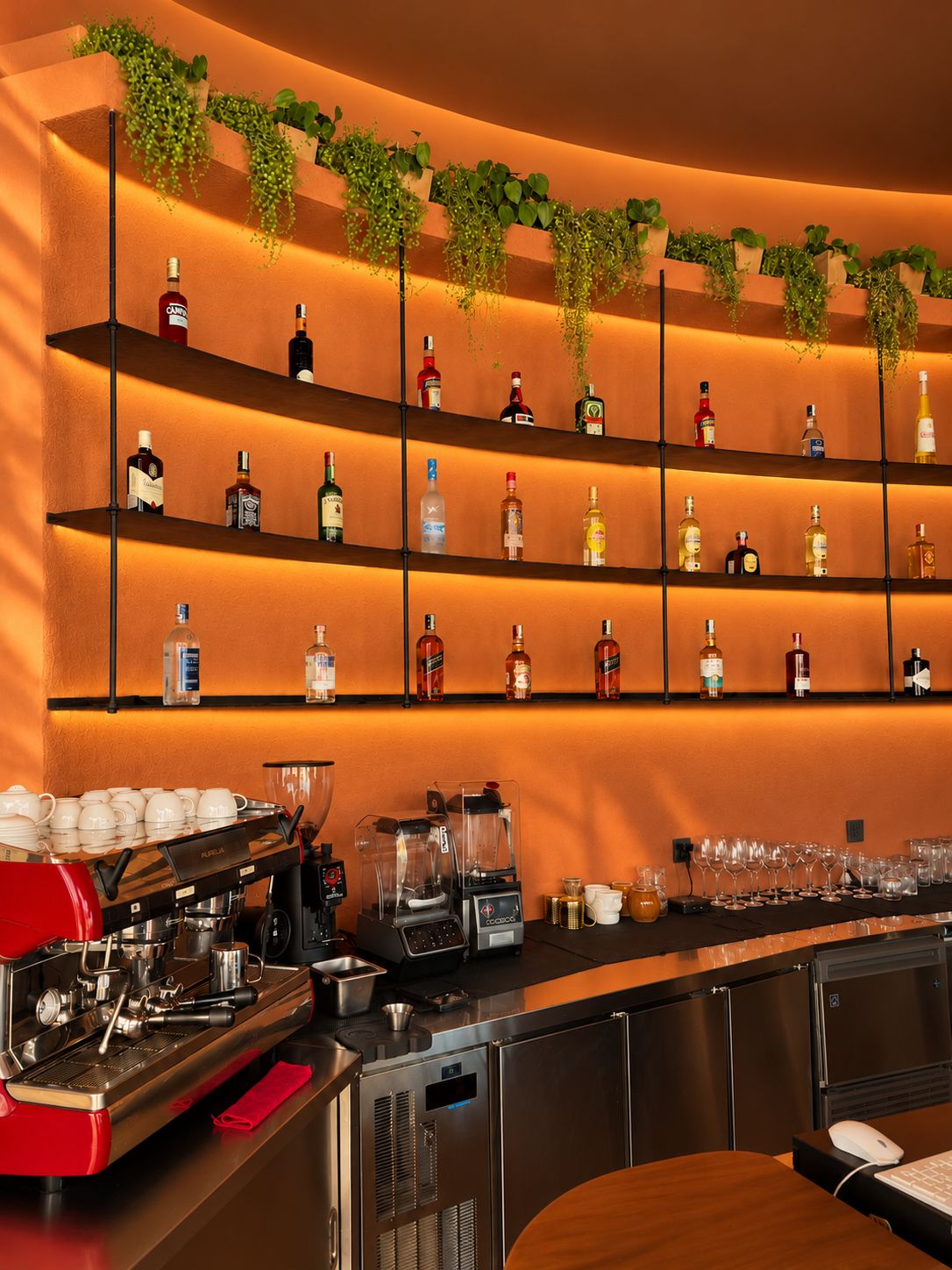 Uma área de bar com prateleiras de garrafas de bebidas, uma máquina de café e um balcão com copos e xícaras.