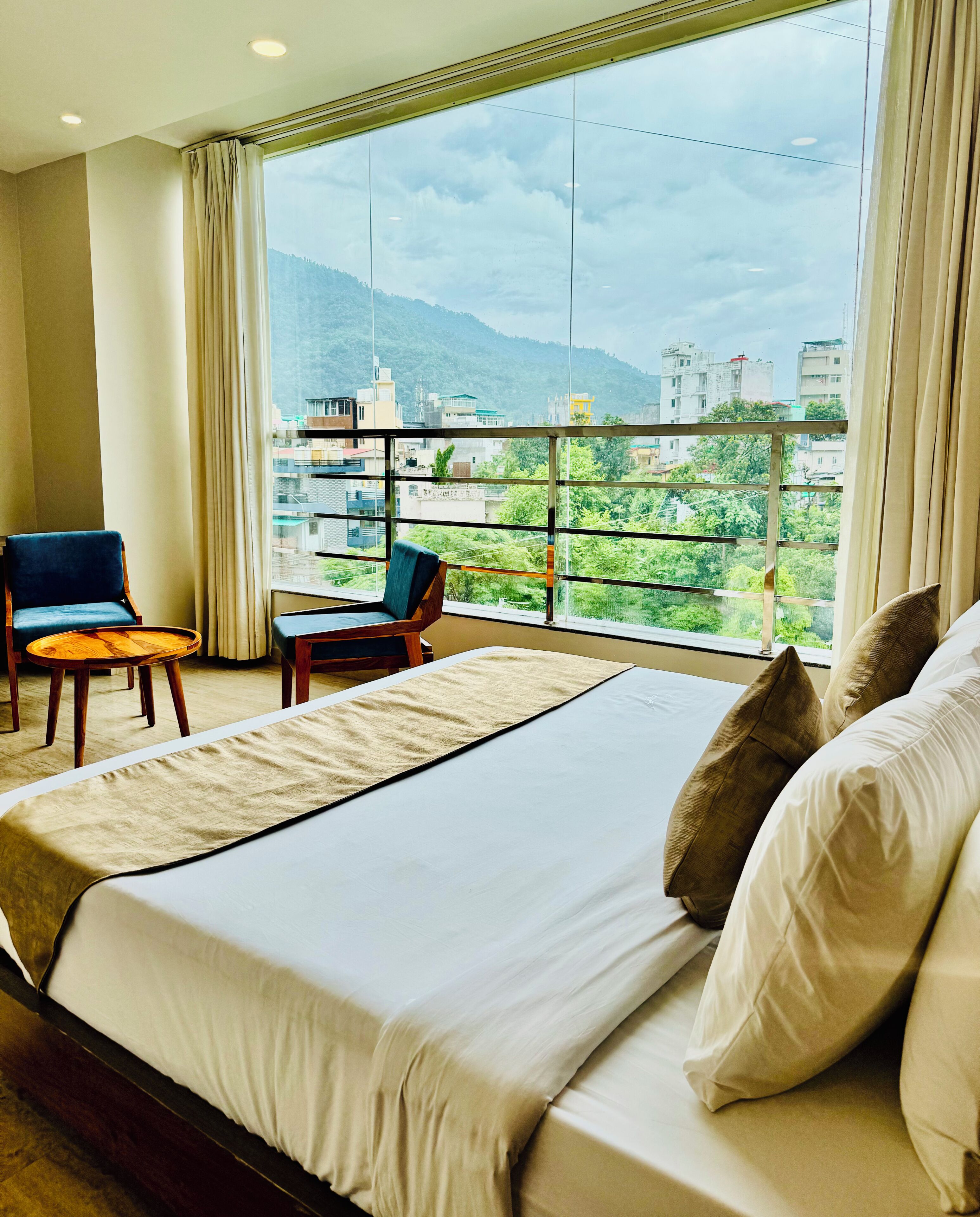 Quarto de hotel com uma cama grande, duas cadeiras e vista para a paisagem urbana através de uma janela ampla.