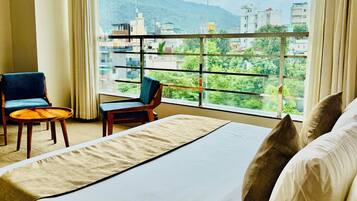 Quarto de hotel com uma cama grande, duas cadeiras e vista para a paisagem urbana através de uma janela ampla.