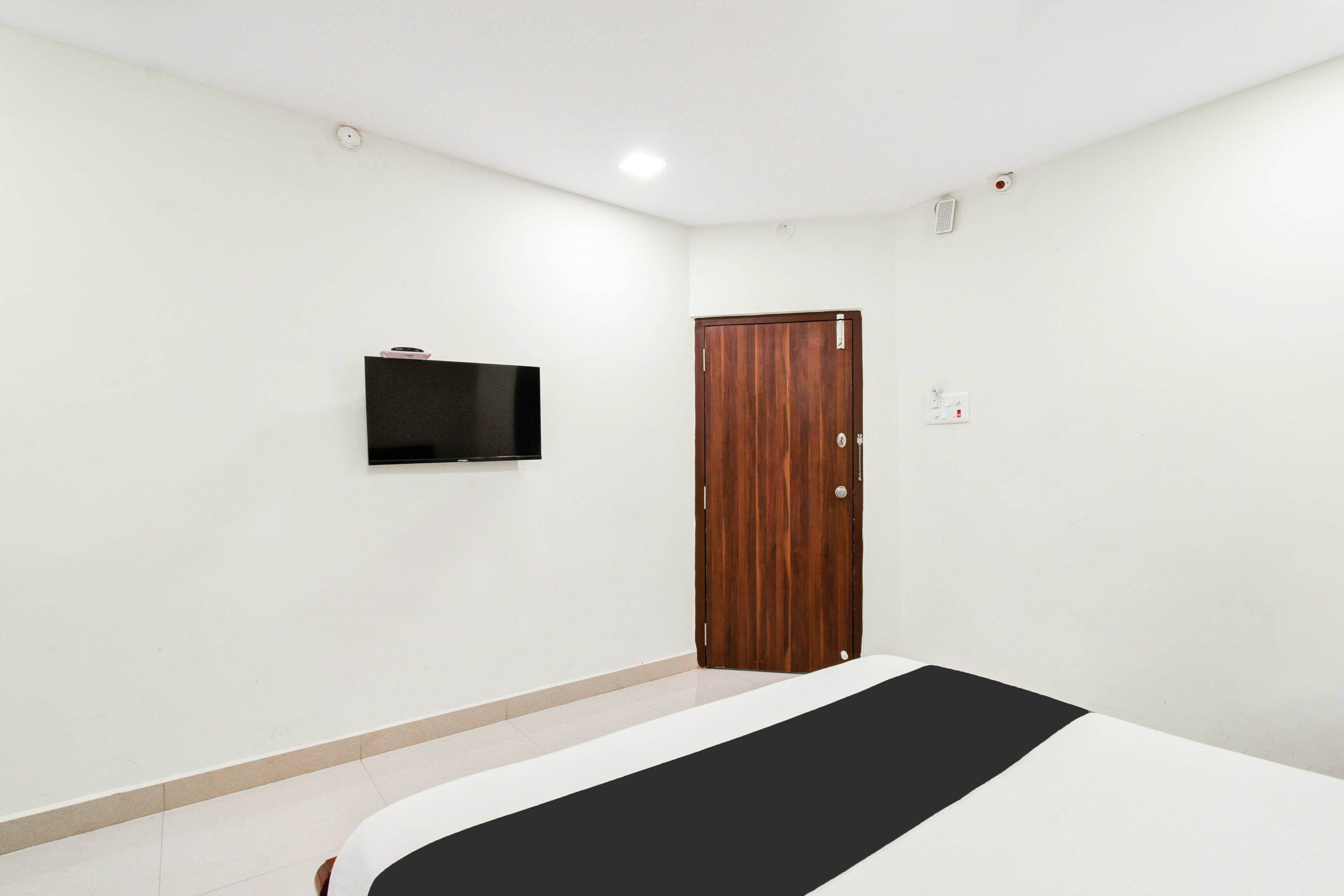 Quarto com cama, porta de madeira, TV fixada na parede e interruptor de luz.