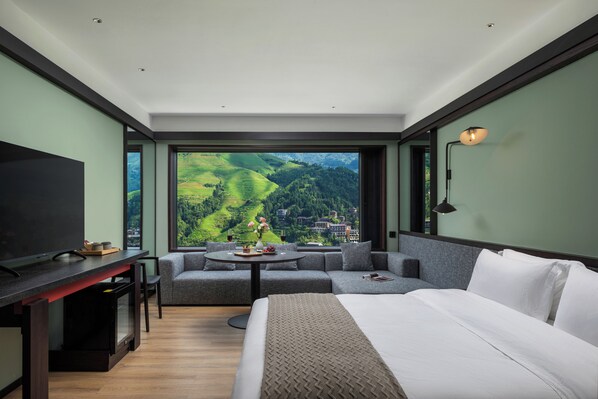 Quarto de hotel moderno com uma cama grande, um sofá, uma mesa de jantar e uma televisão. O quarto possui uma janela ampla com vista para colinas verdes e uma cidade.