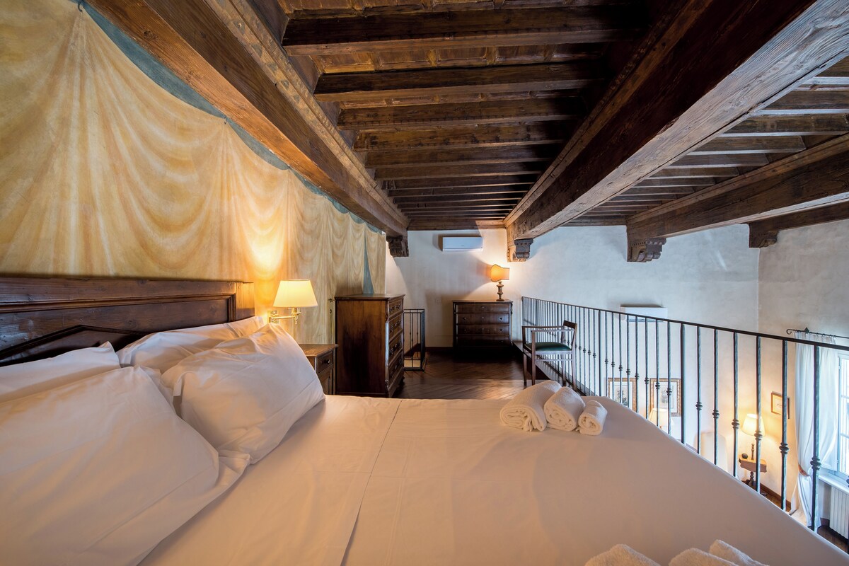 Quarto de hotel com uma cama grande, vigas de madeira, uma varanda com guarda-corpo e uma escada.