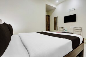 Quarto de hotel com cama, televisão de tela plana na parede, uma cadeira e uma mesa pequena com garrafa e copos.
