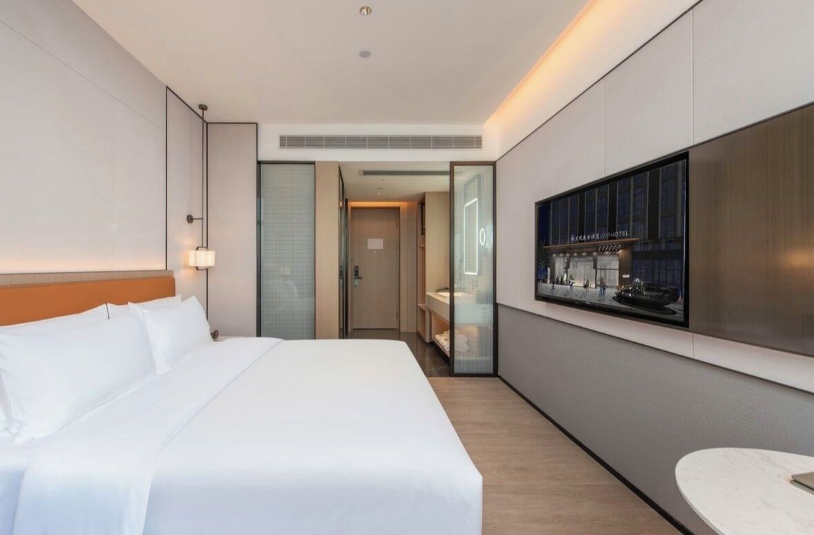 Quarto de hotel com uma cama grande, uma televisão fixada na parede e uma mesa redonda pequena.