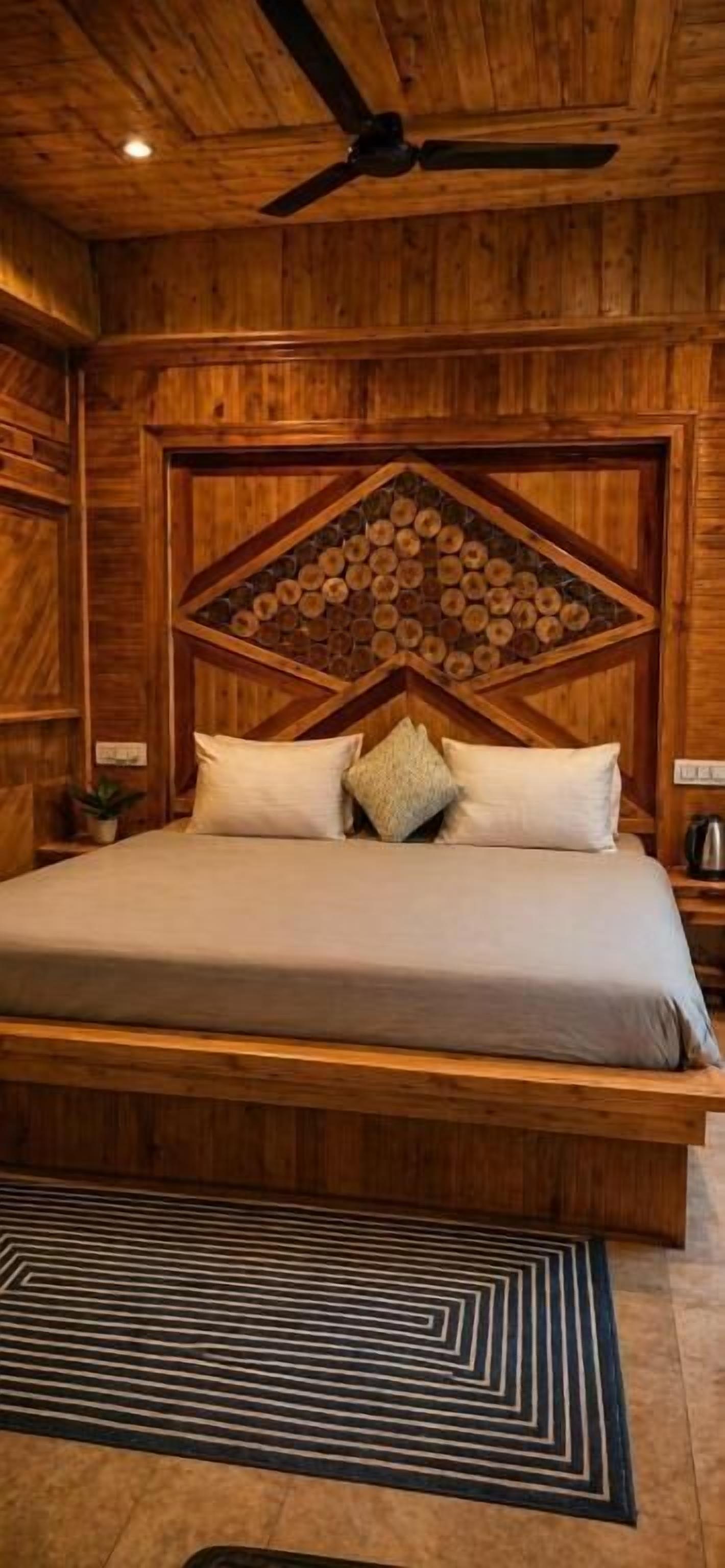 Quarto de madeira com cama, ventilador de teto e cabeceira decorativa.