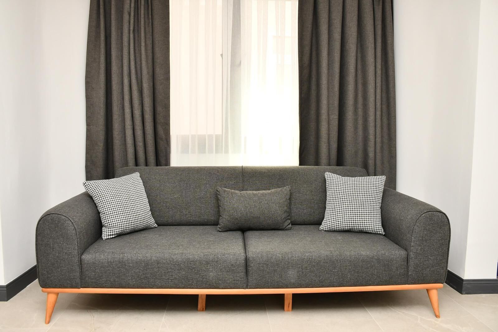 Um sofá cinzento com pernas de madeira e duas almofadas estampadas, posicionado em frente a uma janela com cortinas leves.
