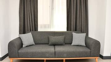 Um sofá cinzento com pernas de madeira e duas almofadas estampadas, posicionado em frente a uma janela com cortinas leves.