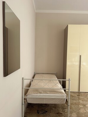 Quarto pequeno com cama de solteiro, estrutura de metal, colchão simples e guarda-roupa com porta.